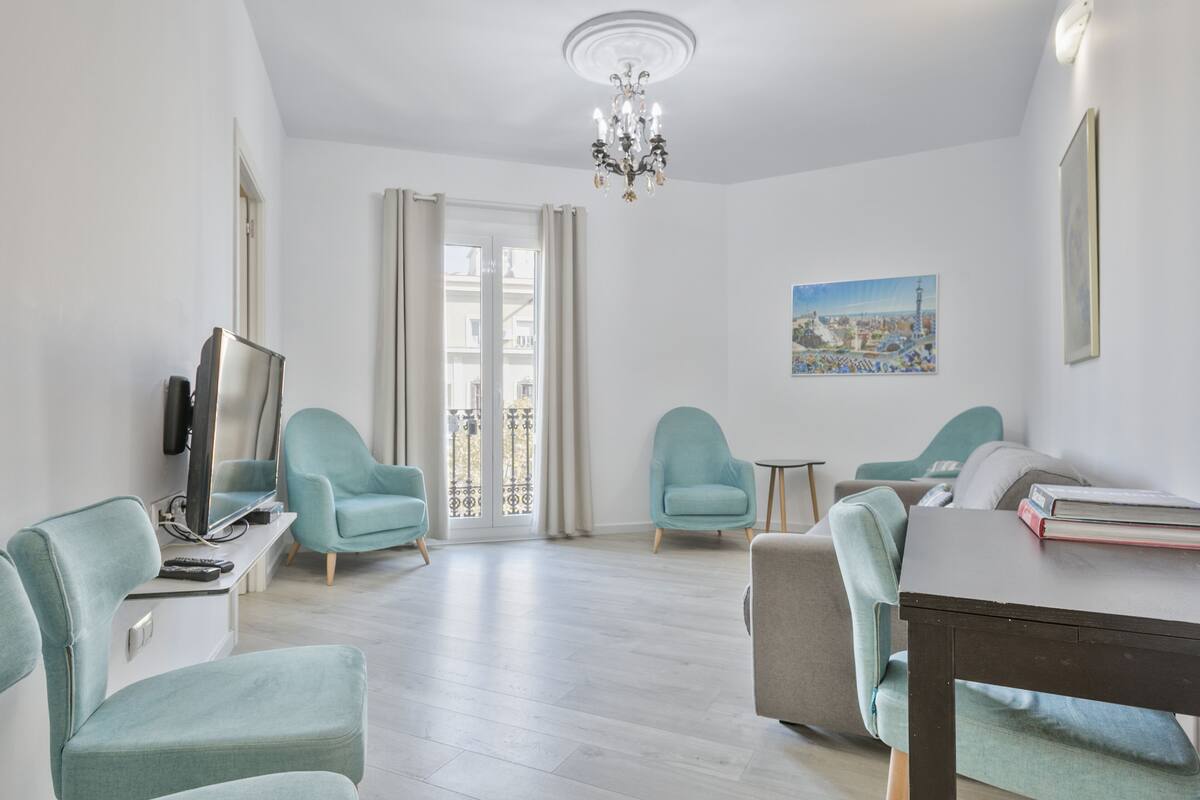 Uma sala de estar moderna com televisão de tela plana, dois poltronas azul-esverdeadas, um sofá cinza, uma mesa de jantar e um lustre.