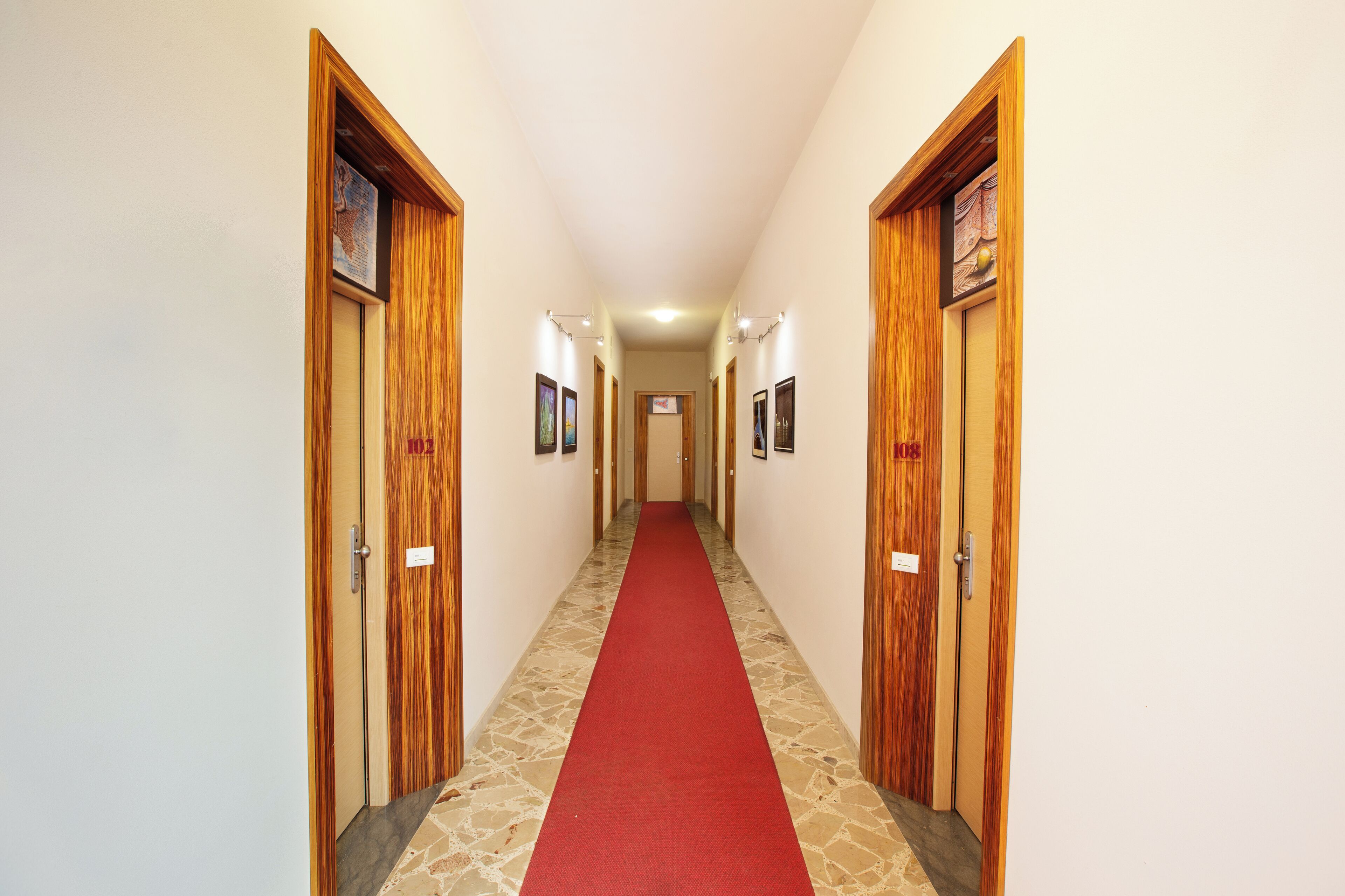 Corredor com portas de madeira, tapete vermelho e quadros nas paredes.