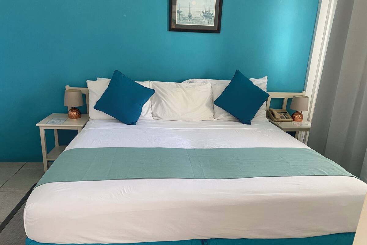 Cama bem arrumada com roupa de cama branca e azul turquesa, dois travesseiros azuis e um cobertor azul turquesa. Há duas mesas de cabeceira com abajures e um quadro na parede.