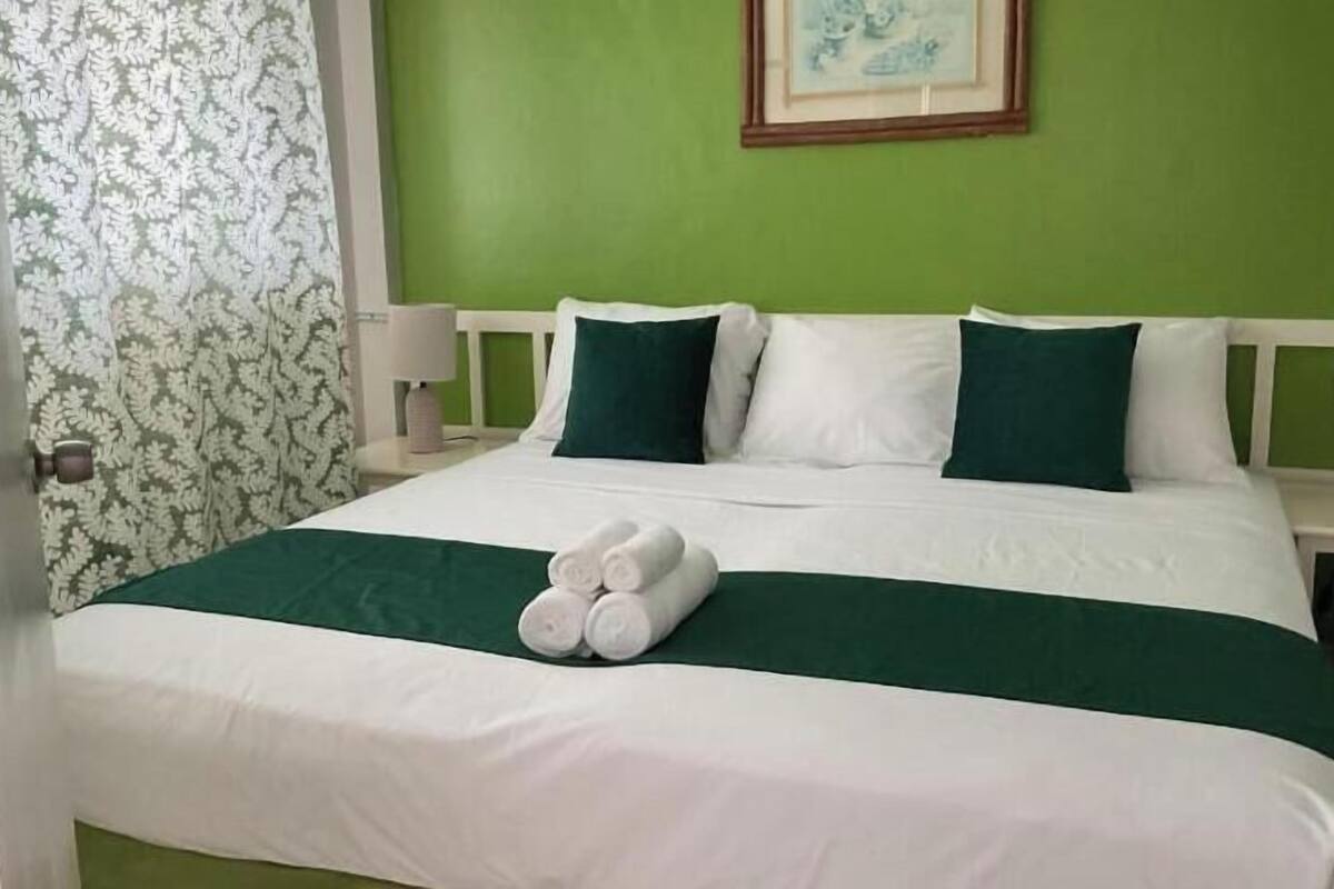 Cama bem arrumada com lençóis brancos e verdes, um par de toalhas enroladas e um quadro na parede.