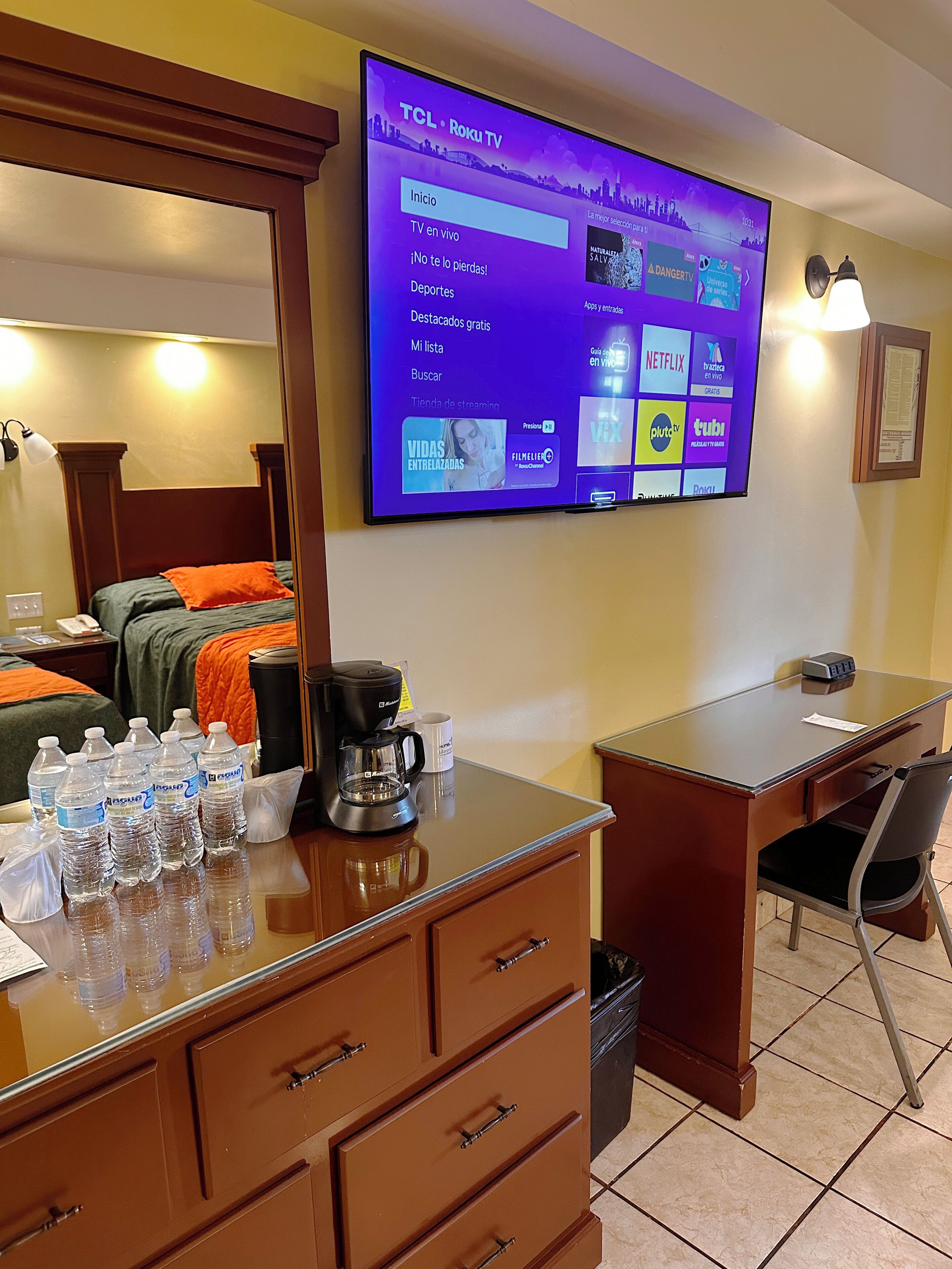 Quarto de hotel com televisão de tela plana exibindo a interface de um serviço de streaming, cômoda de madeira com garrafas de água e máquina de café, cama com roupa de cama laranja e escrivaninha com cadeira.