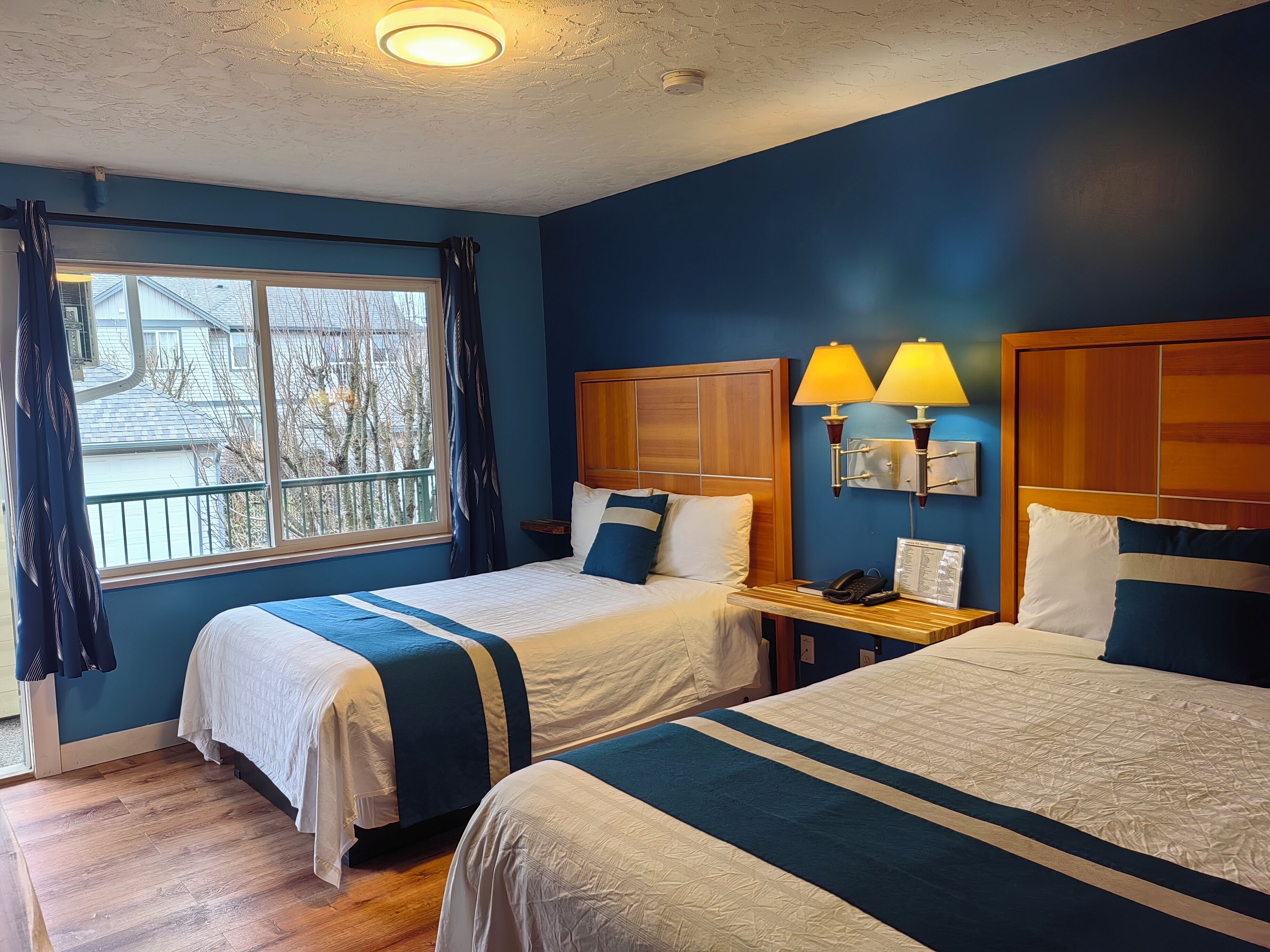Quarto de hotel com duas camas, uma janela com cortinas, uma cabeceira de madeira e uma mesa de cabeceira com abajur e telefone.