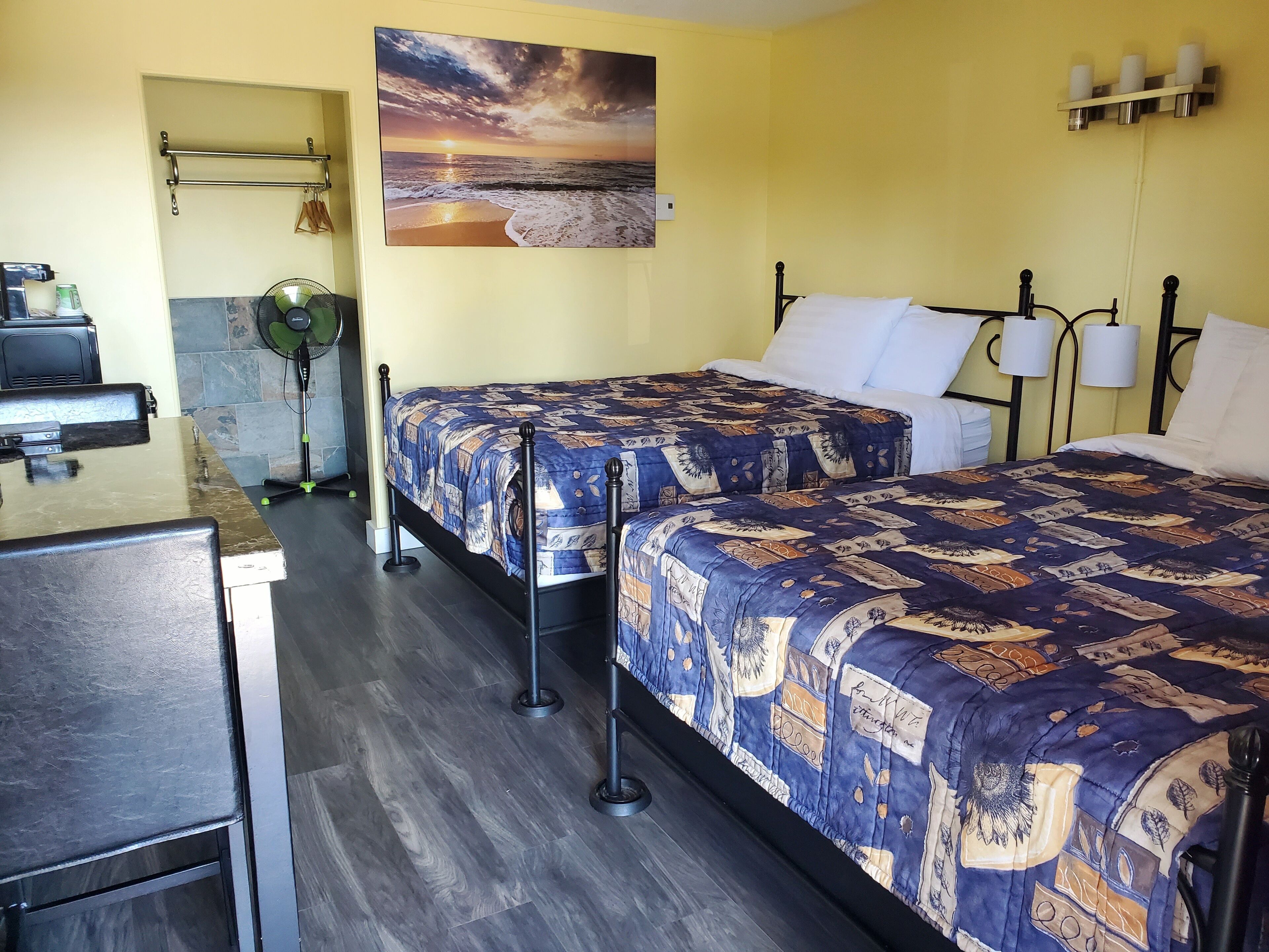 Quarto de hotel com duas camas, uma grande pintura de um pôr do sol, uma pequena escrivaninha e um ventilador.