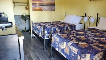 Quarto de hotel com duas camas, uma grande pintura de um pôr do sol, uma pequena escrivaninha e um ventilador.