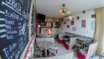 Um café com menu em lousa listando diversas cervejas e seus preços, mobiliário de madeira e um espaço de estar aconchegante.