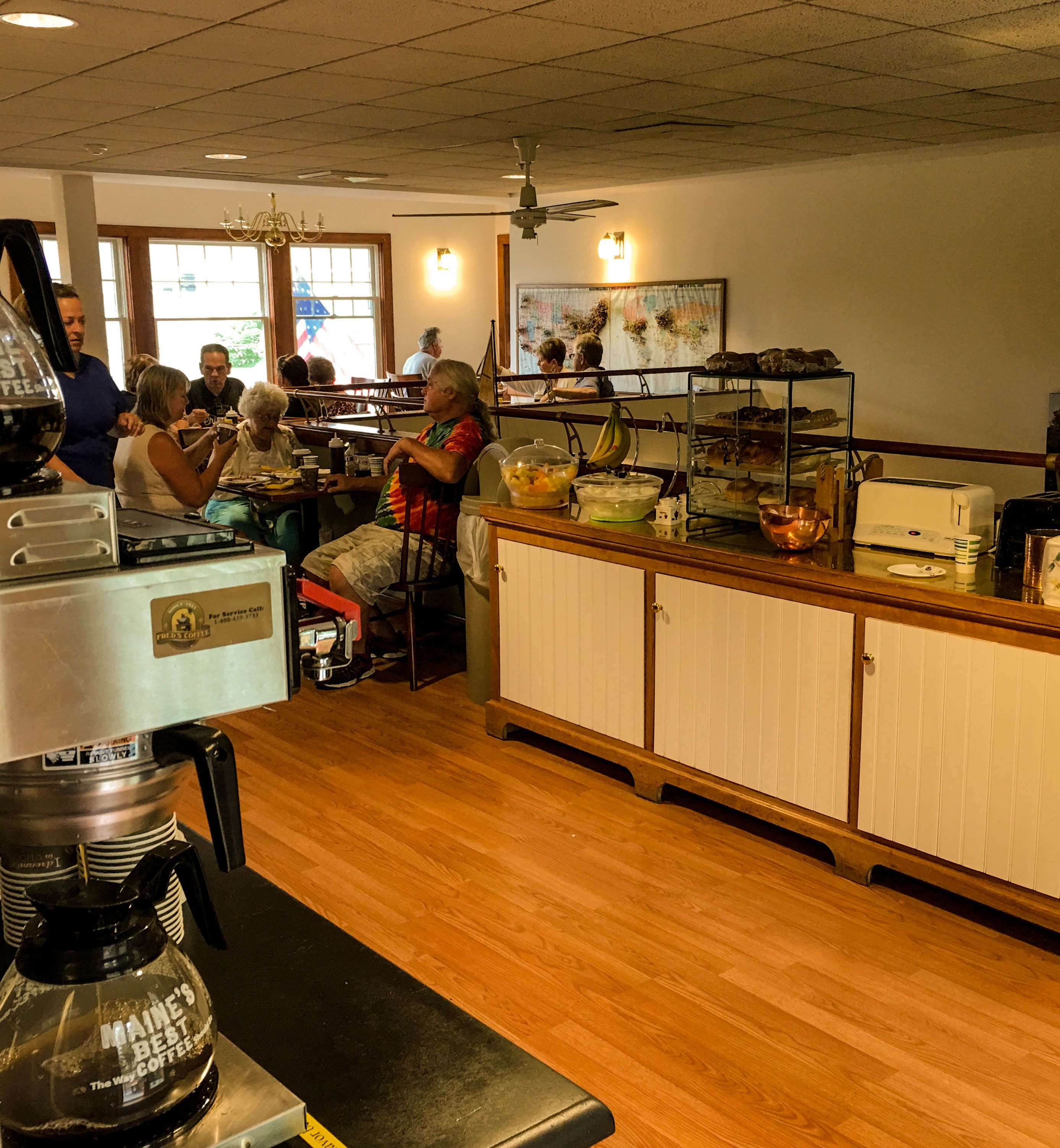 Interior de café com piso de madeira, máquina de café, balcão com vitrine e clientes sentados em mesas.