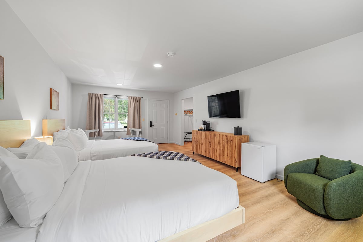 Quarto de hotel moderno com duas camas, uma poltrona verde, uma televisão e uma escrivaninha de madeira.