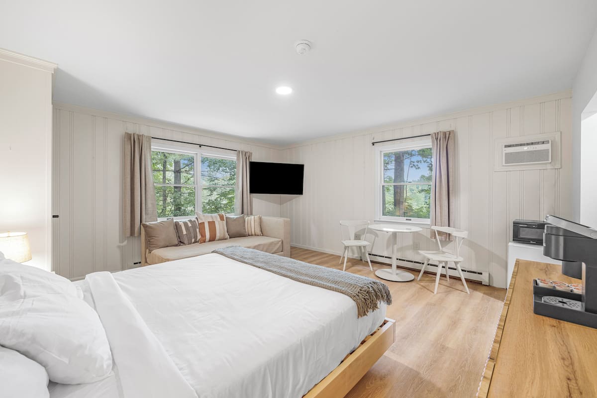 Quarto de hotel moderno com uma cama grande, uma escrivaninha com televisão de tela plana, uma pequena mesa de jantar e uma janela com cortinas.