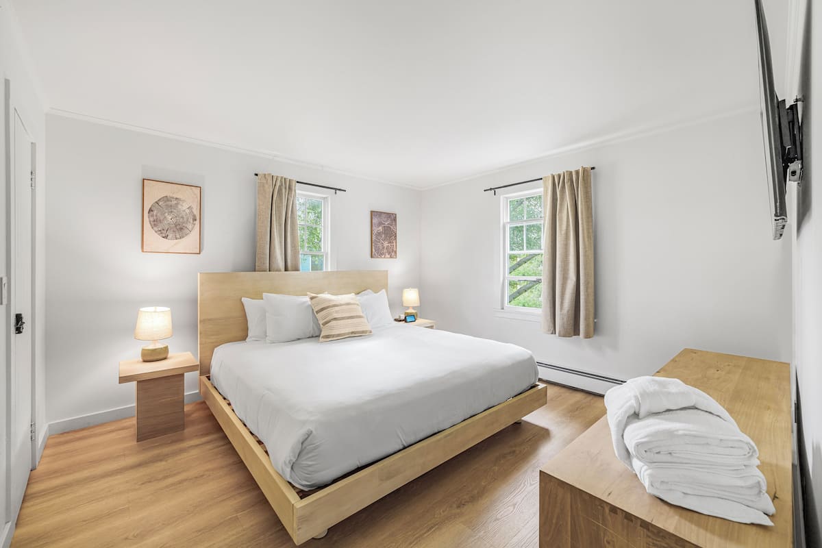 Quarto de hotel moderno com cama de madeira, duas mesas de cabeceira com luminárias, vista para área verde pelas janelas e toalha dobrada sobre um banco de madeira.