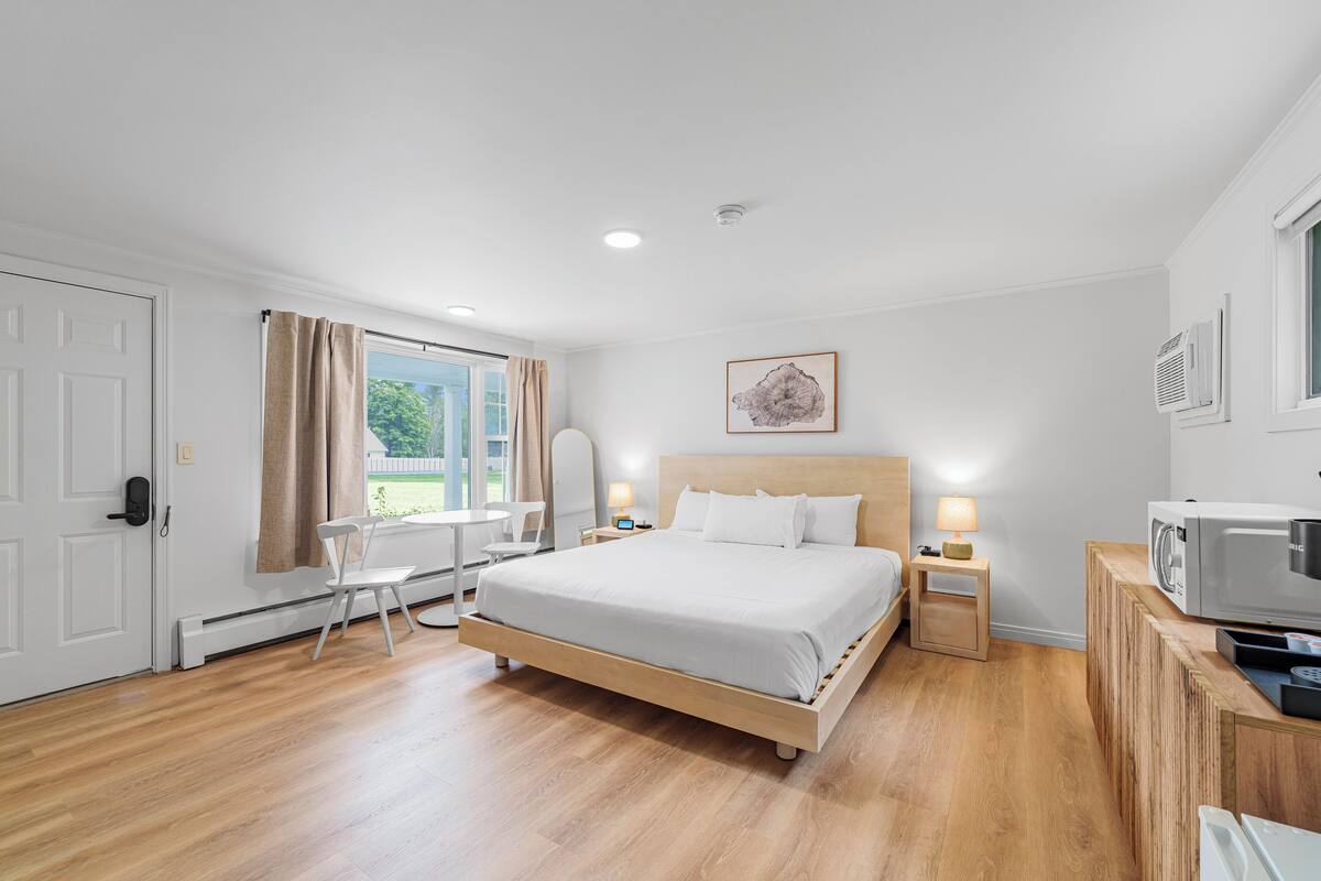 Quarto de hotel moderno com piso de madeira, uma cama grande com roupa de cama branca, uma escrivaninha com cadeira, uma mesinha de cabeceira com abajur e uma janela com cortinas.