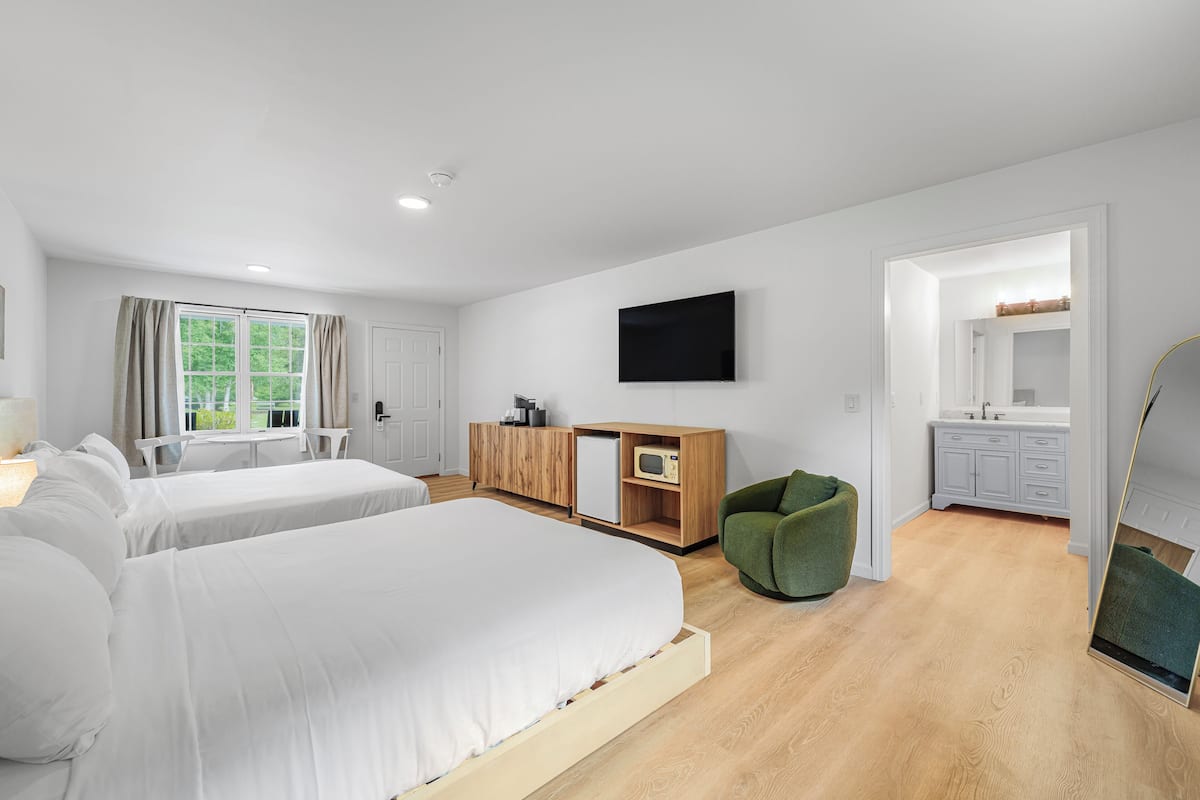 Quarto de hotel moderno com duas camas, uma TV, um armário de madeira, uma poltrona verde e um banheiro visível por uma porta aberta.