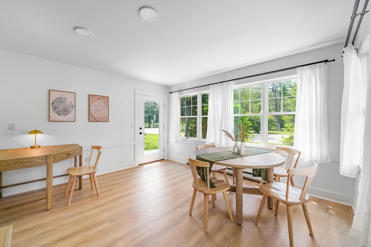 Um quarto espaçoso com uma mesa de jantar de madeira e cadeiras, uma escrivaninha com luminária, e grandes janelas com cortinas.