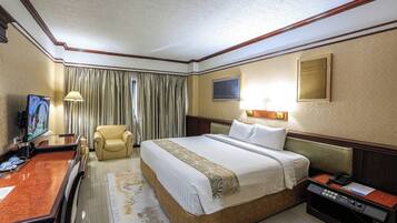 Quarto de hotel com uma cama, uma escrivaninha com televisão, uma cadeira e uma mesinha de cabeceira com telefone.