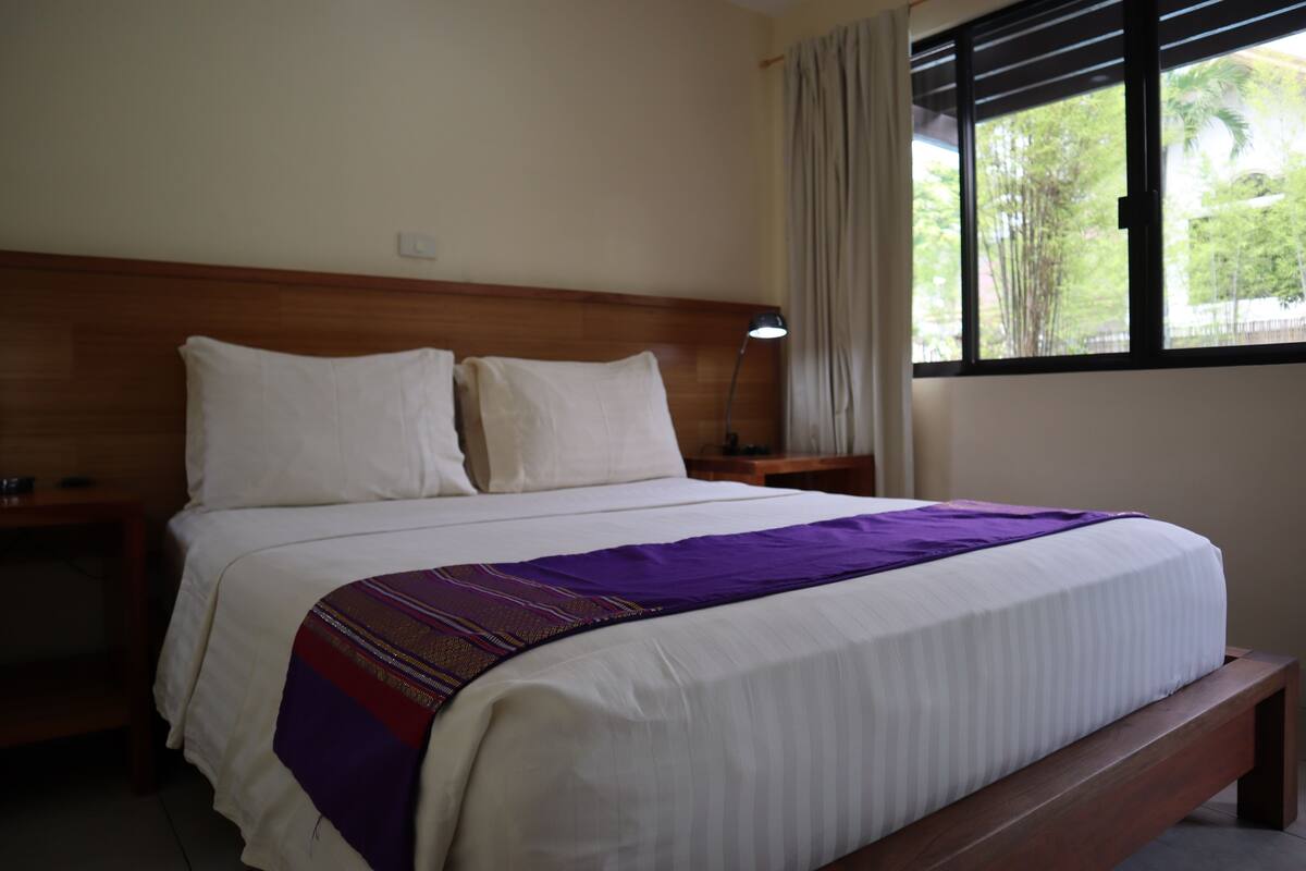 Cama bem arrumada com cabeceira de madeira, mesa de cabeceira com abajur e janela com cortinas.