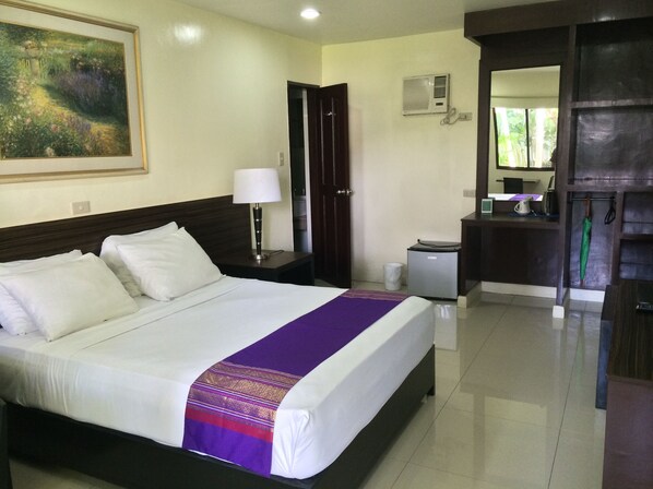 Quarto de hotel com cama, mesa de cabeceira, luminária, lixeira, escrivaninha com computador e espelho.