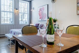 Uma mesa de jantar com uma garrafa de vinho e dois copos, uma janela com vista para árvores e uma cama com um cobertor xadrez.