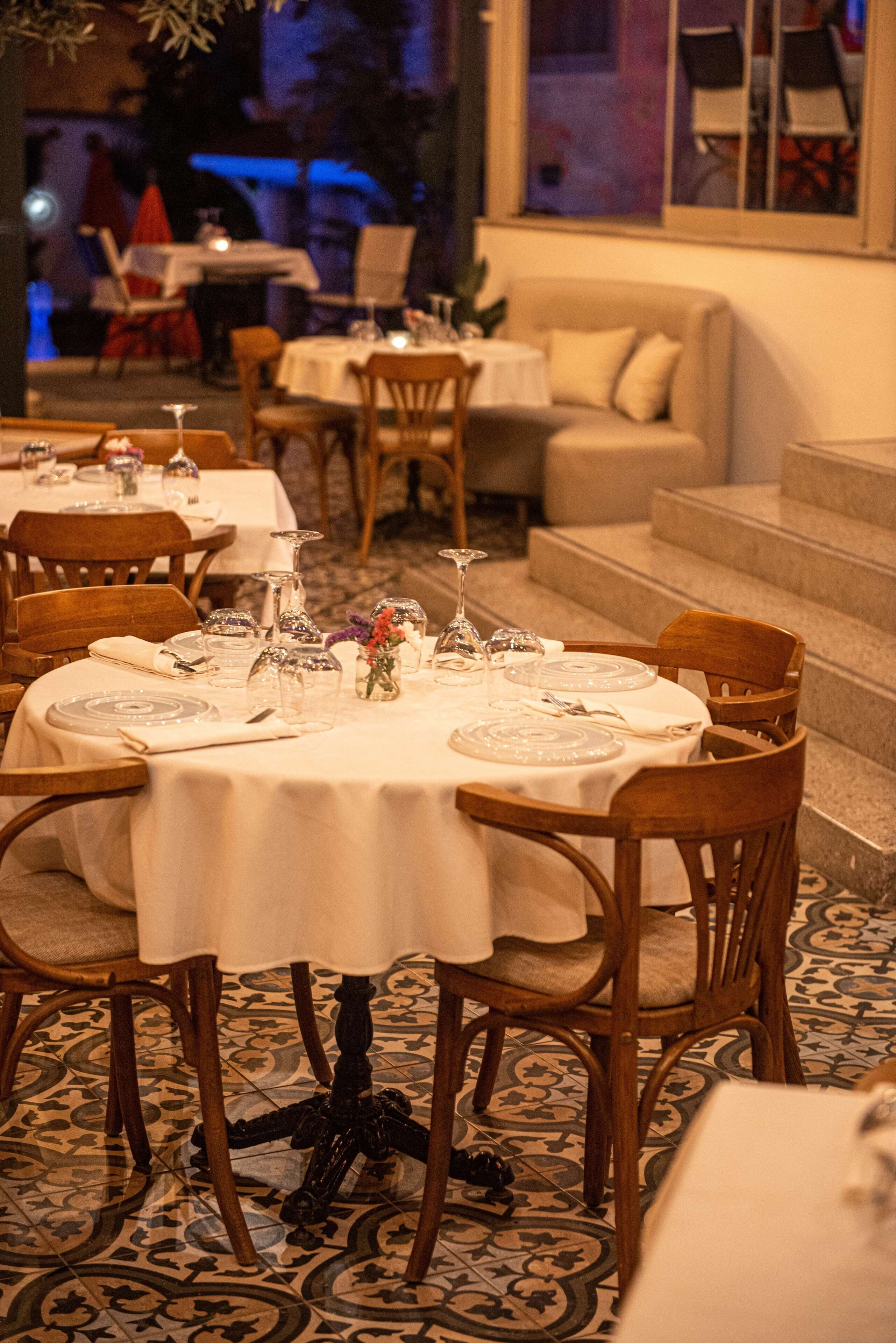 Um restaurante com mesas redondas preparadas para refeições, cadeiras de madeira e um chão estampado.