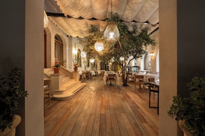 Um espaço de refeições interno com piso de madeira, mesas, cadeiras, luminárias pendentes e plantas em vasos.