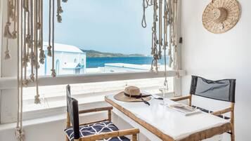 Um quarto com mesa e cadeiras de madeira, luminária de palha no teto e vista para o mar, além de uma parede branca com um enfeite de tear.
