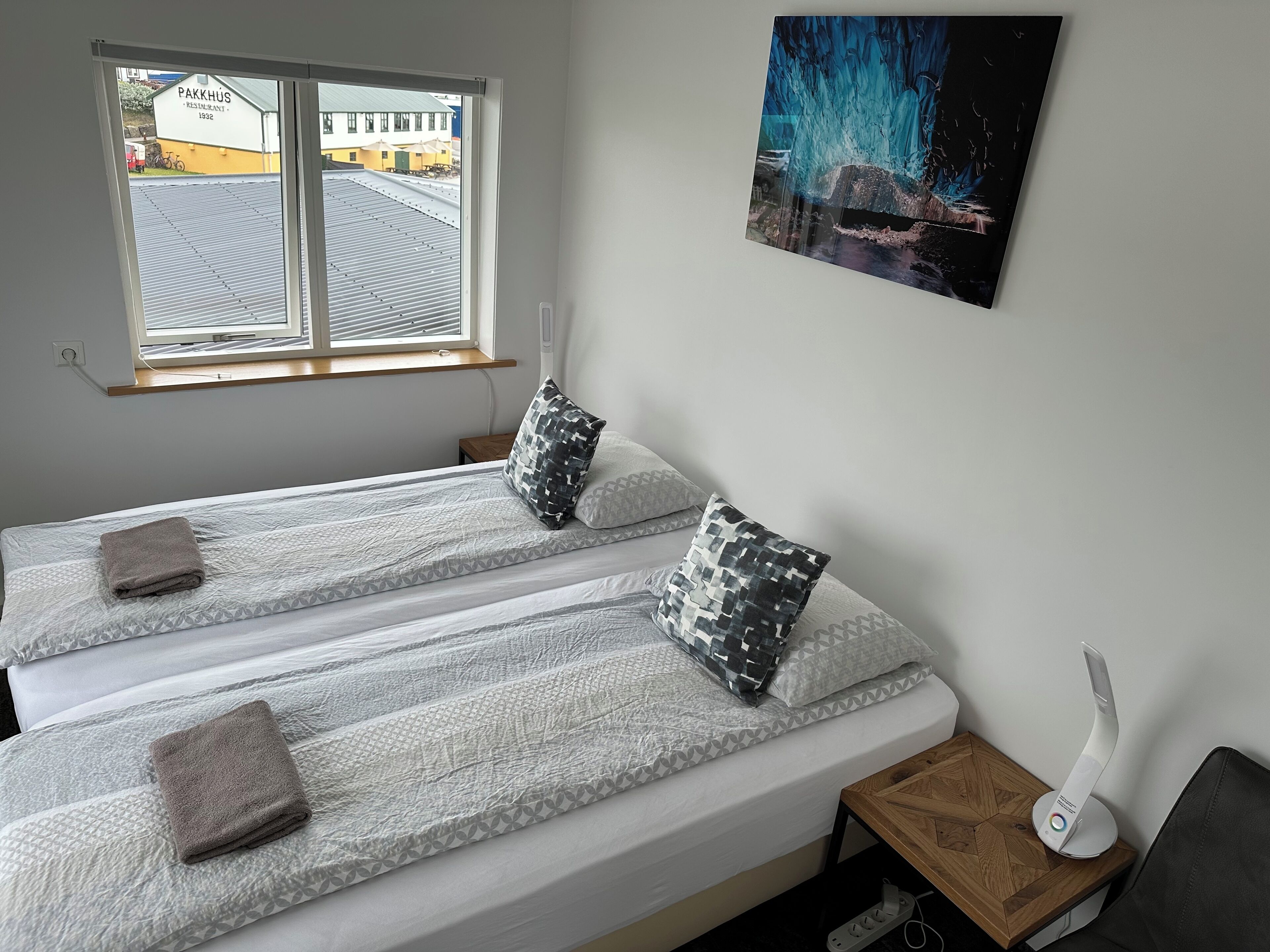Uma cama de casal com roupa de cama estampada, uma pequena mesa de cabeceira com abajur e vista para um edifício na janela.