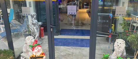 Entrada de hotel com porta de vidro, placa de boas-vindas e estátuas decorativas na parte externa.