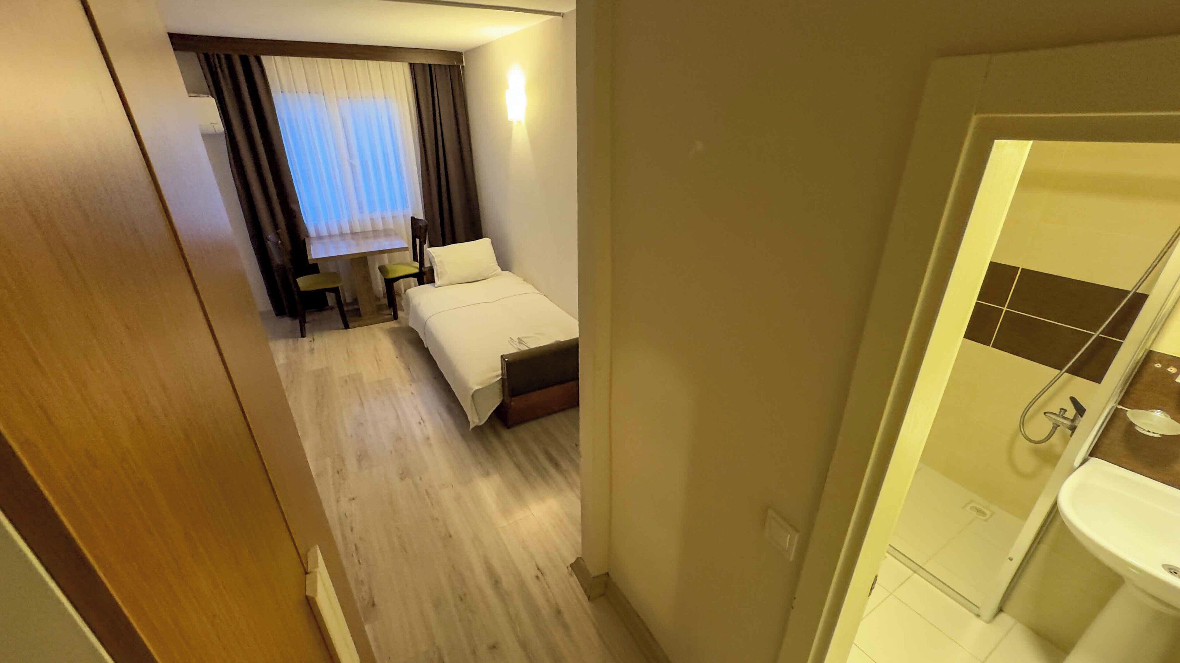 Quarto de hotel com cama, escrivaninha e cadeira. Inclui banheiro com pia e chuveiro.