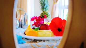 Uma travessa de fruta com uma flor vermelha, uma abacaxi e um copo de sumo de laranja sobre uma mesa com toalha de mesa florida.