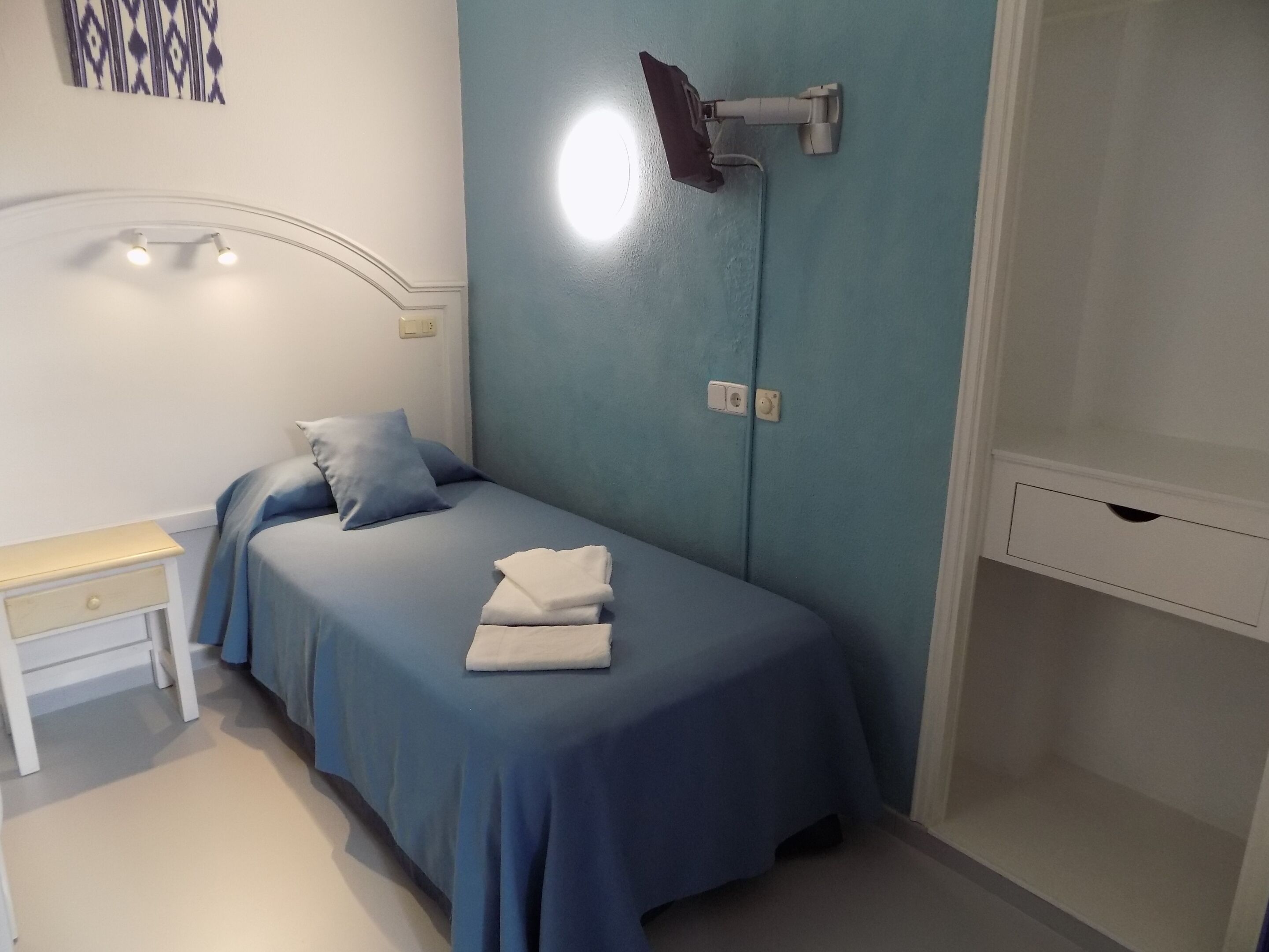 Quarto com uma cama de solteiro, mesa de cabeceira, luminária na parede e um pequeno armário branco.