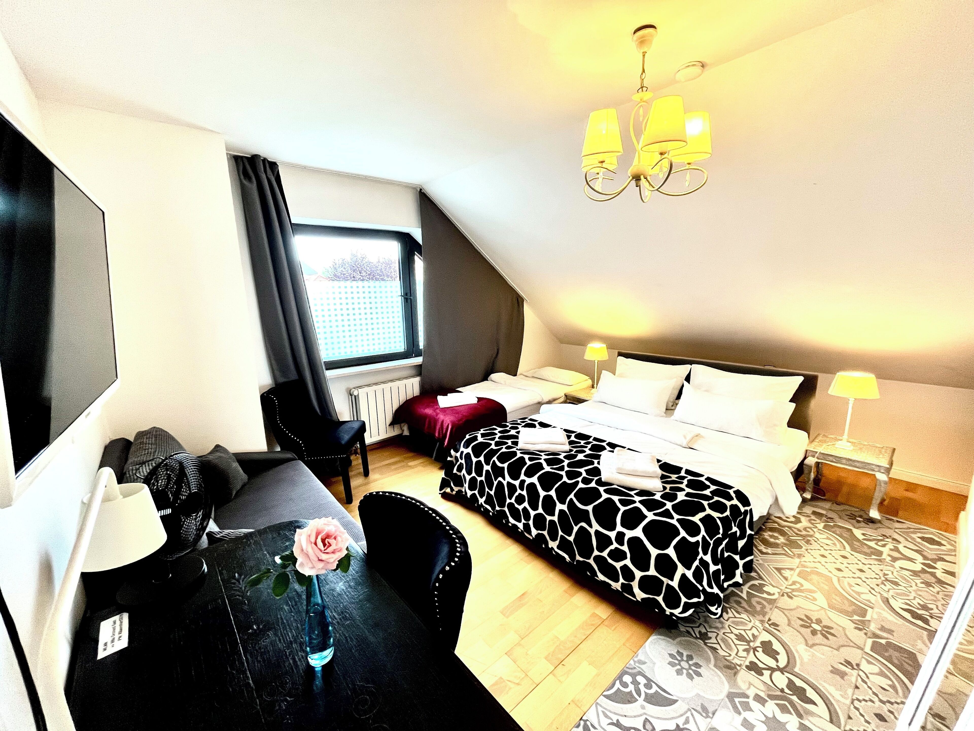 Quarto de hotel com cama, escrivaninha, cadeira, pequena mesa com um vaso de flores e janela com cortinas.