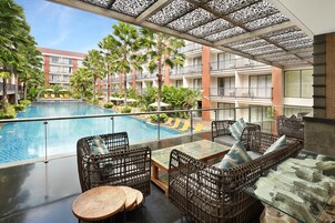 Uma área à beira da piscina com mobiliário de vime e uma mesa de vidro, com vista para uma piscina e um edifício com varandas.