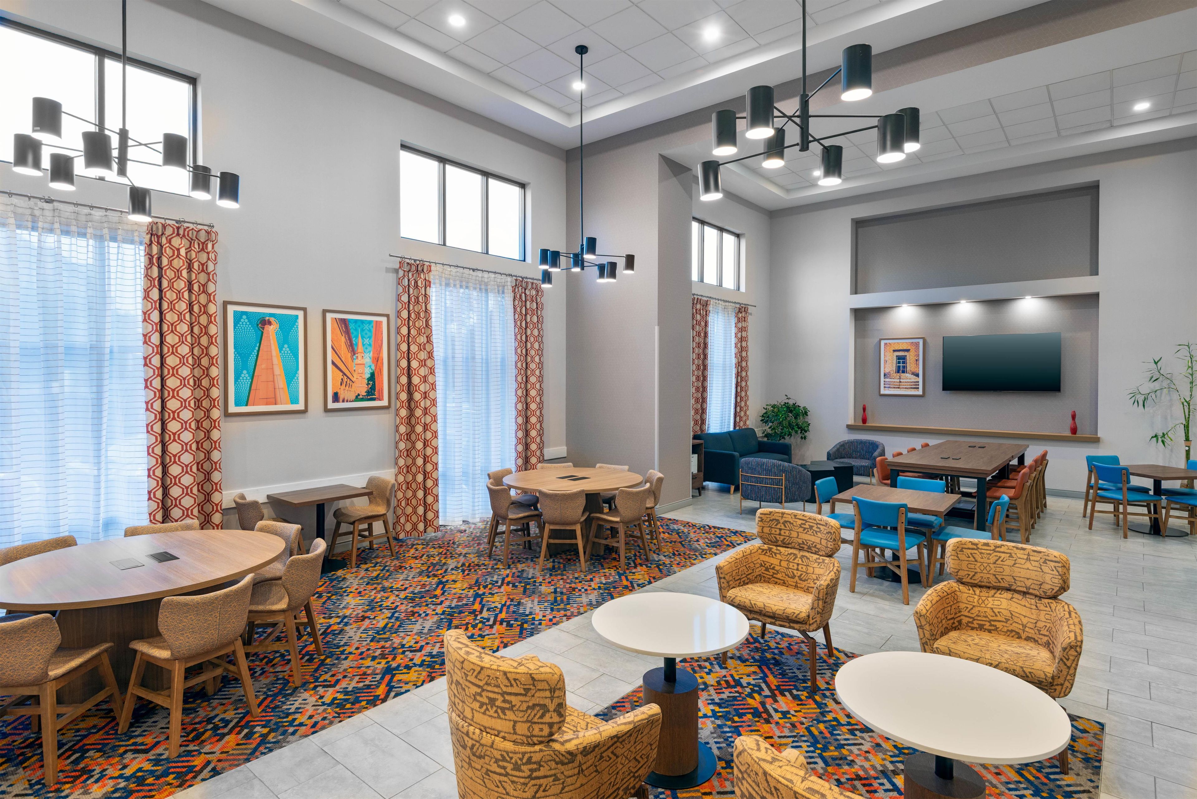 Um saguão de hotel com uma grande mesa de reuniões, várias mesas menores, assentos confortáveis, obras de arte nas paredes e uma TV de tela plana fixada na parede.