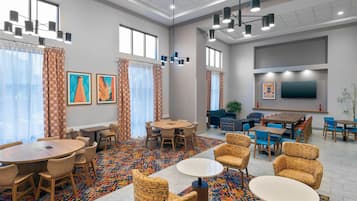 Um saguão de hotel com uma grande mesa de reuniões, várias mesas menores, assentos confortáveis, obras de arte nas paredes e uma TV de tela plana fixada na parede.