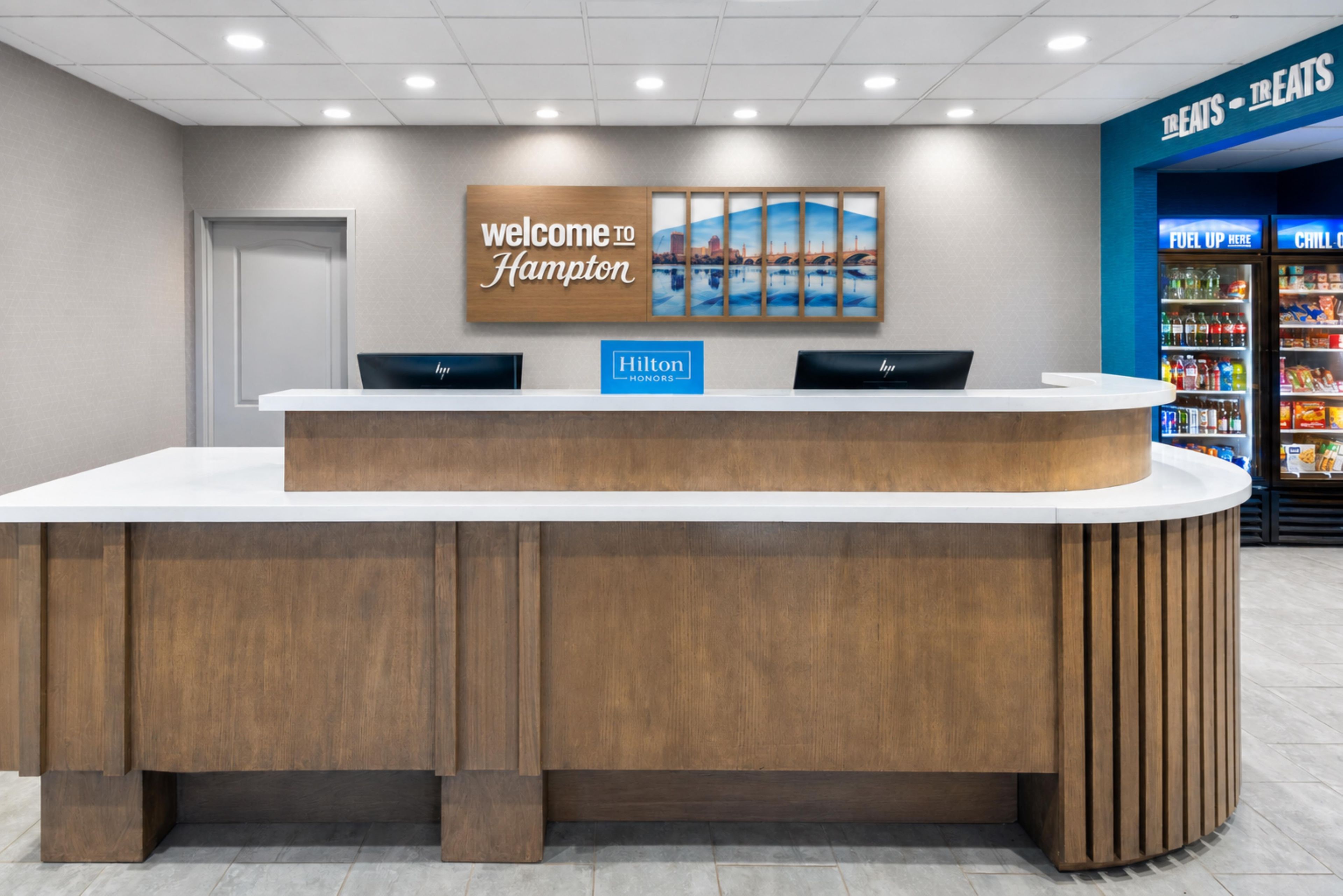 Uma mesa de recepção com a placa 'Welcome to Hampton', uma credencial 'Hilton Honors' e uma geladeira 'Fuel Up Here'.