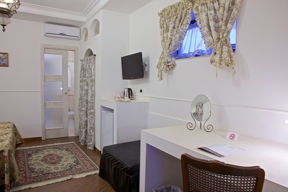 Quarto de hotel com escrivaninha, cadeira, televisão, janela com cortinas, mesinha com chaleira e tapete.