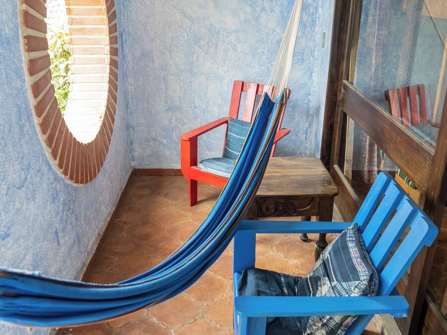 Uma rede estendida entre duas cadeiras azuis sobre um chão de azulejos, com um arco de tijolos e uma janela ao fundo.