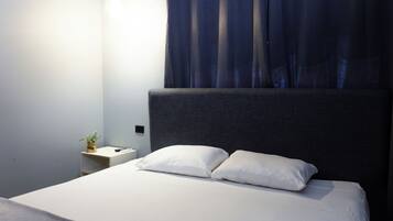 Quarto de hotel com uma cama grande, uma mesa de cabeceira, um abajur e uma janela com cortinas.