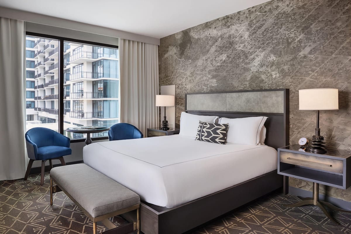 Quarto de hotel com uma cama grande, duas mesas de cabeceira, uma escrivaninha, uma cadeira e vista para um edifício alto.