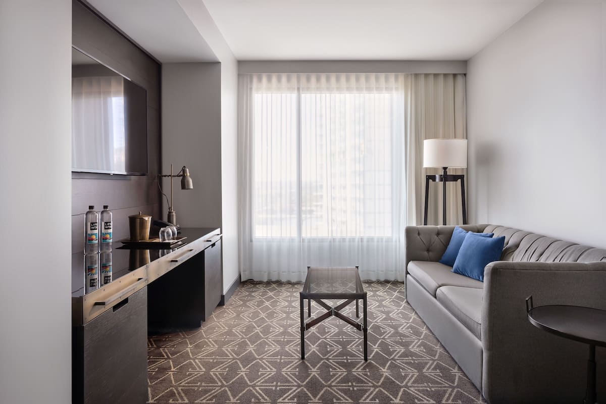 Quarto de hotel compacto com cozinha compacta, sofá, mesa pequena e uma janela grande com cortinas translúcidas.