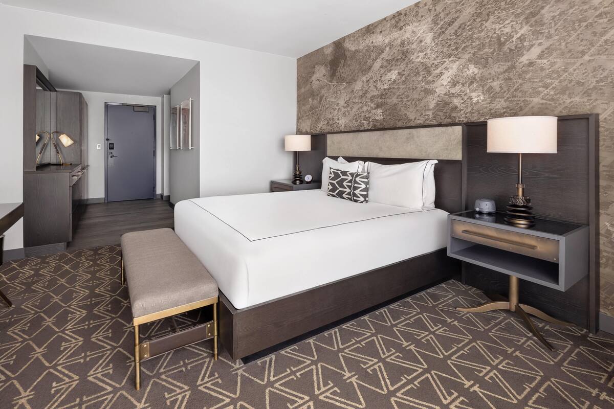 Quarto de hotel moderno com uma cama grande, mesas de cabeceira, um banco e uma parede com textura.