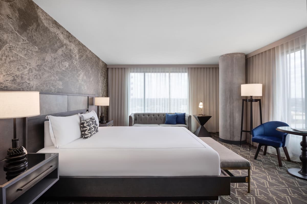 Quarto de hotel com uma cama grande, um sofá, uma poltrona, uma mesa de trabalho e uma luminária.