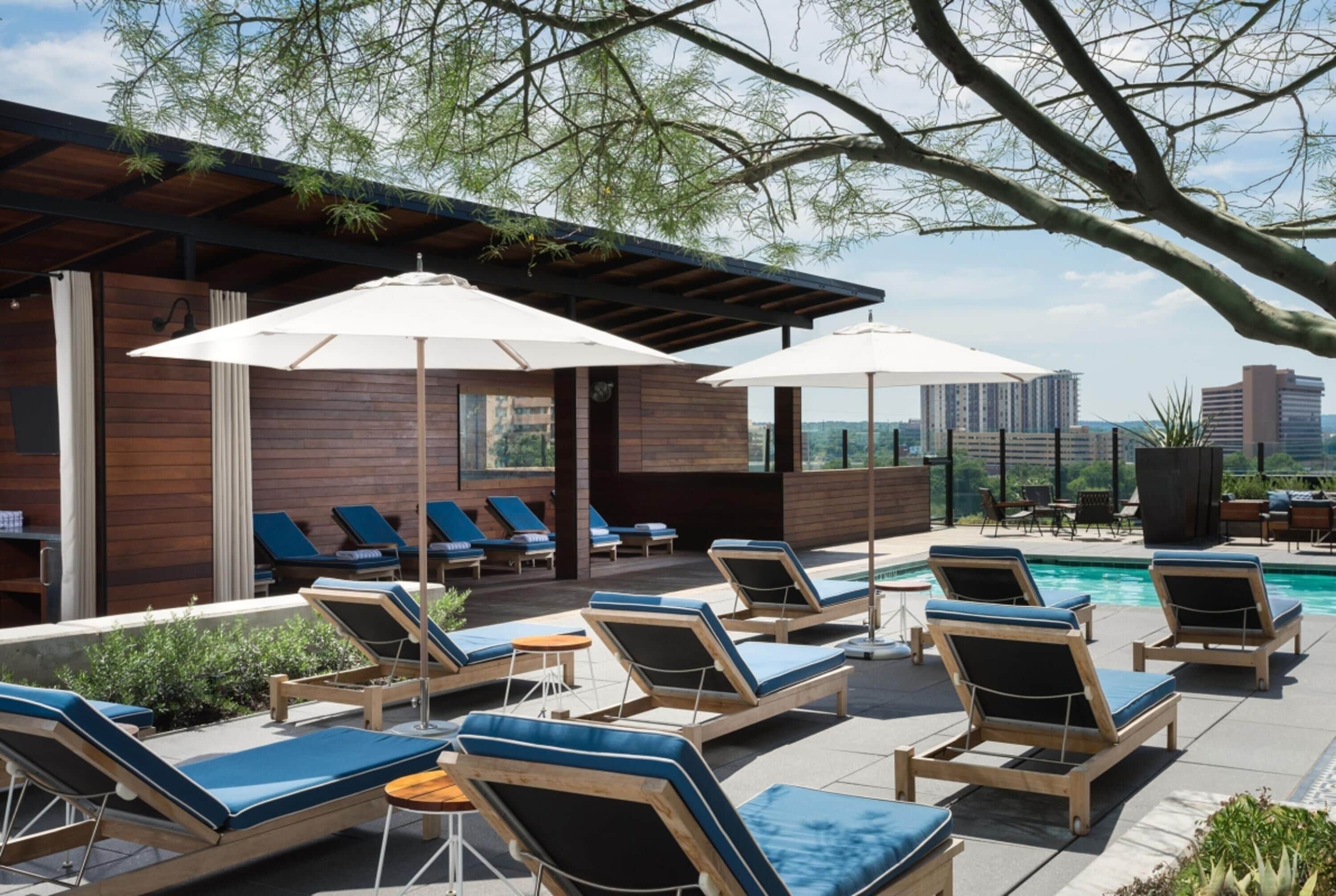 Uma área de piscina no terraço, com espreguiçadeiras e guarda-sóis, deck de madeira, e vista para o horizonte da cidade.