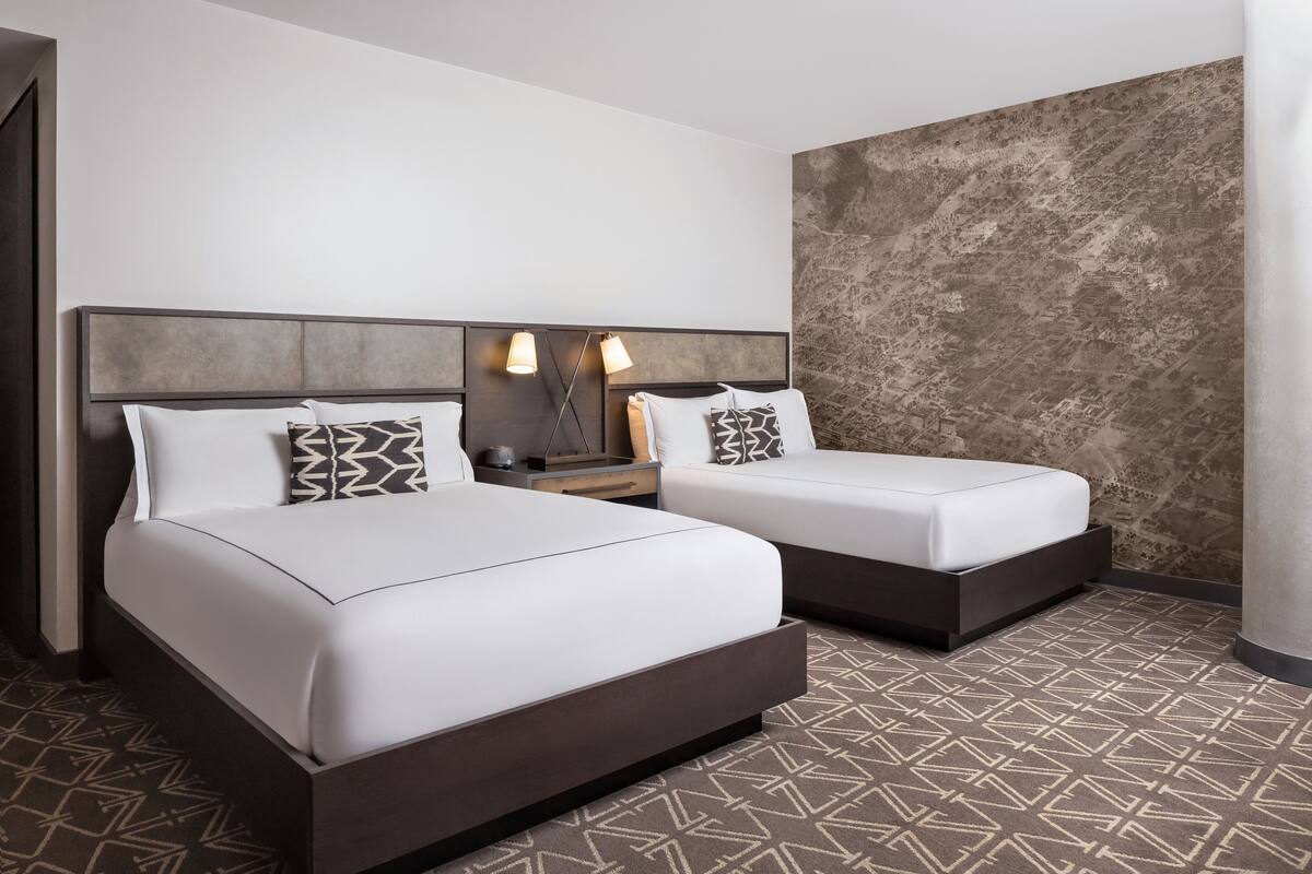 Quarto de hotel com duas camas, cabeceira, mesas de cabeceira e parede com textura.