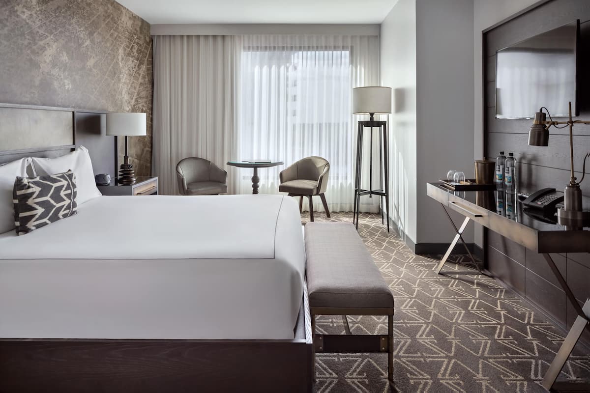 Quarto de hotel com uma cama grande, uma escrivaninha com luminária, uma cadeira, uma mesinha e uma TV fixada na parede.