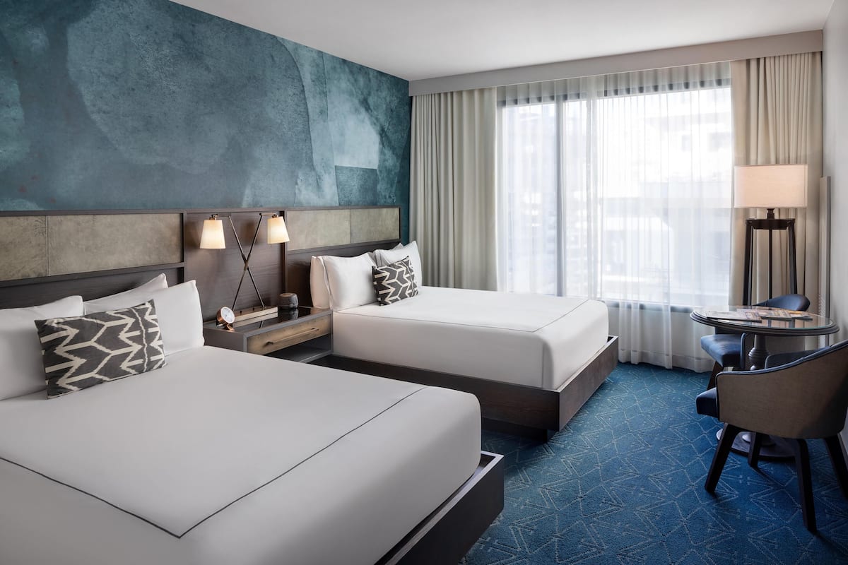 Quarto de hotel com duas camas, uma escrivaninha, uma cadeira, um abajur e uma janela grande com cortinas.