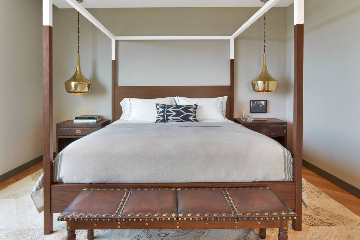Uma cama de dossel com estrutura de madeira, duas mesas de cabeceira e um banquinho ao pé da cama.