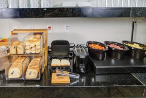 Disposição de buffet com pães, torradas e diversos recipientes de comida.