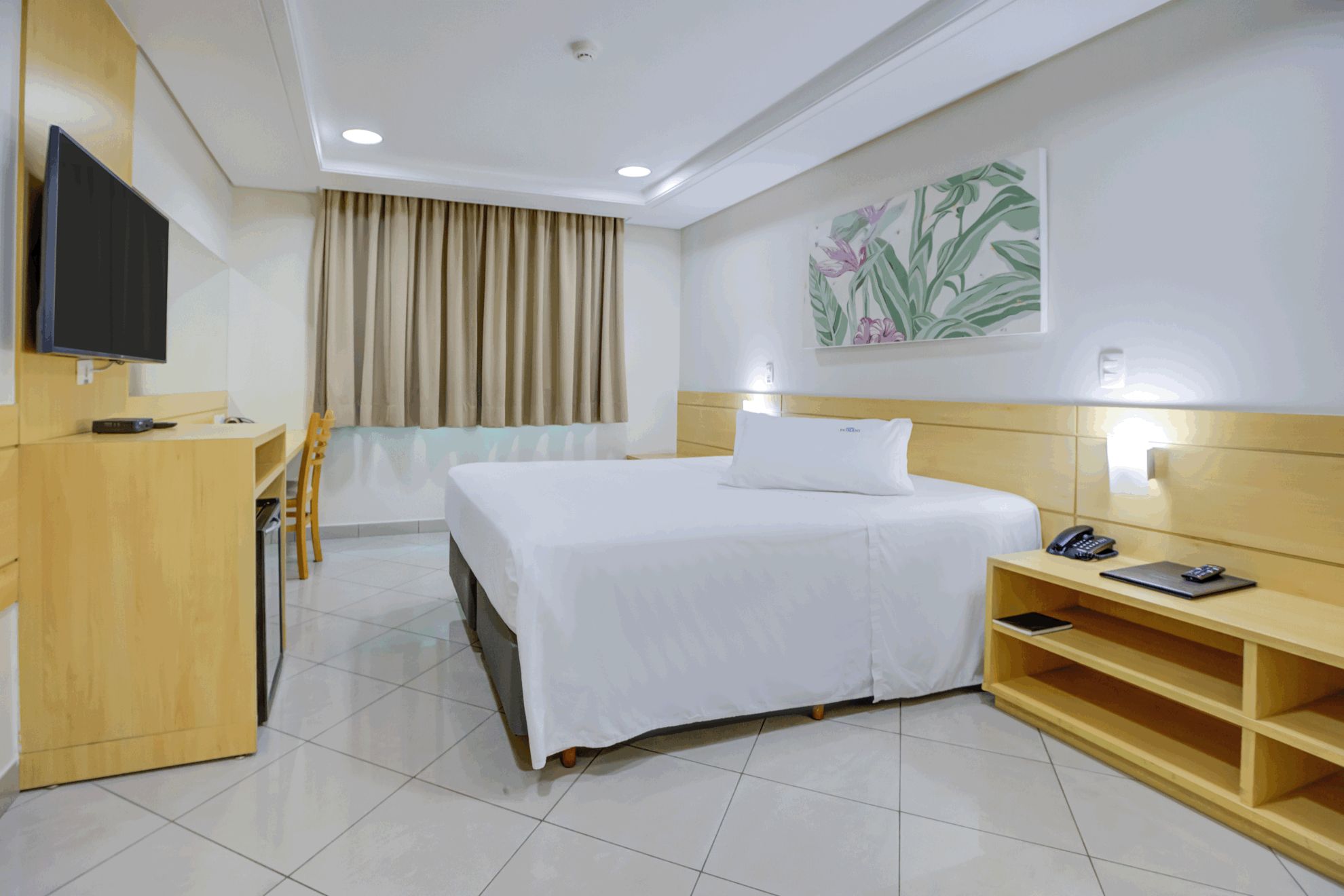 Quarto de hotel com cama, escrivaninha, cadeira, televisão, quadro e telefone.