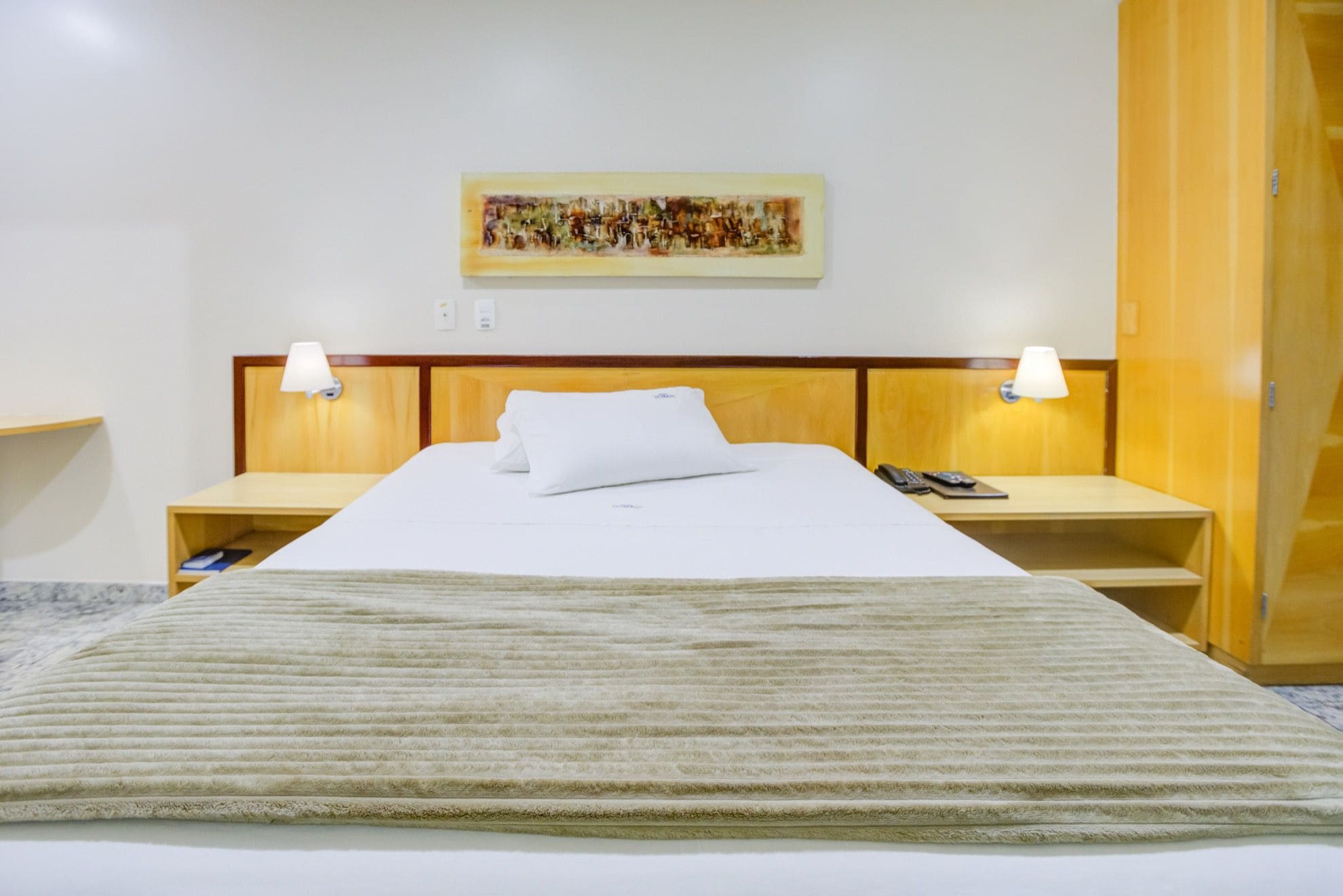 Quarto de hotel com uma cama grande, duas mesas de cabeceira, uma cabeceira, um quadro na parede e uma porta.