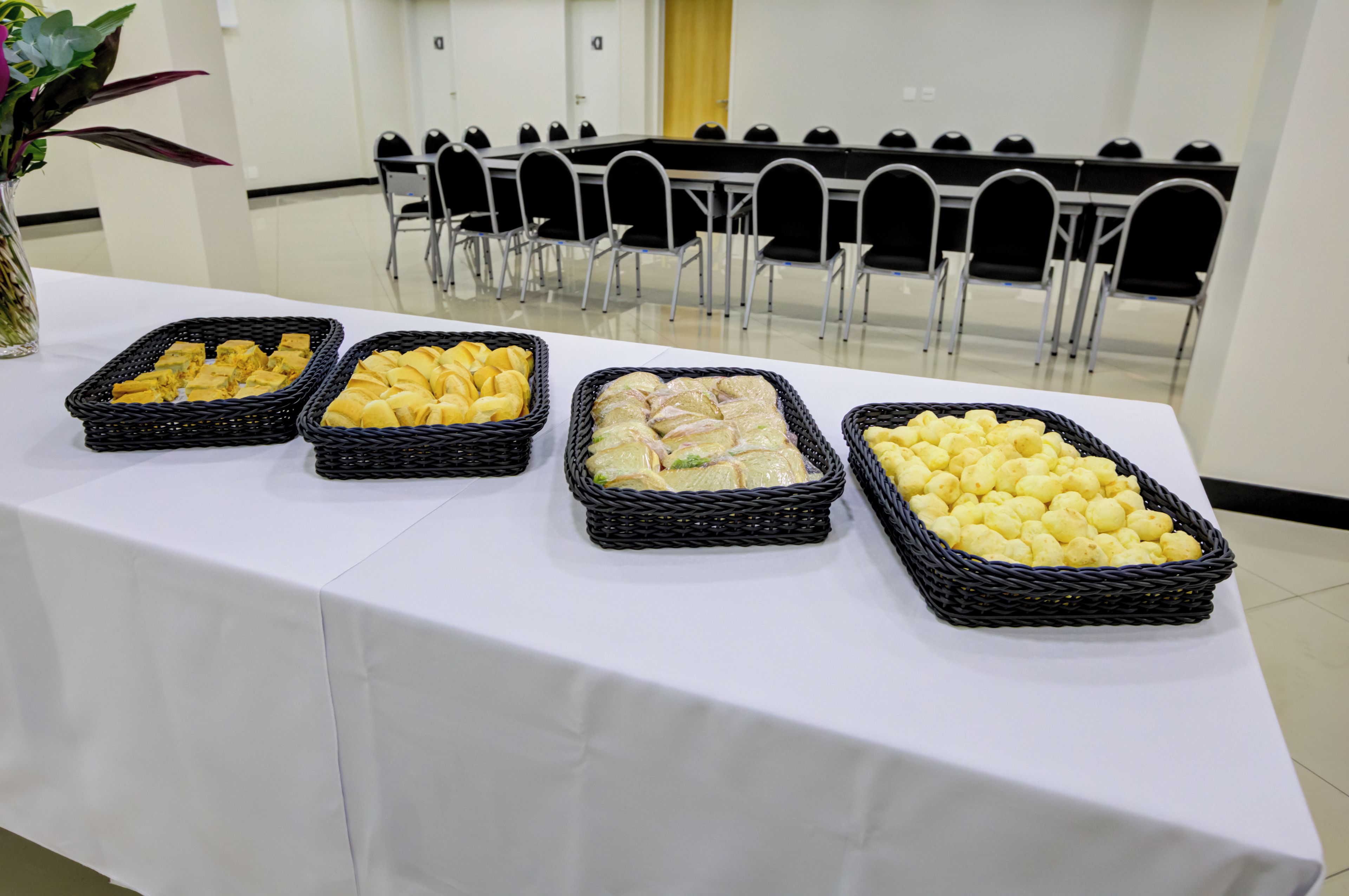Mesa de buffet com quatro cestos de alimentos, toalha de mesa branca e fileiras de cadeiras ao fundo.
