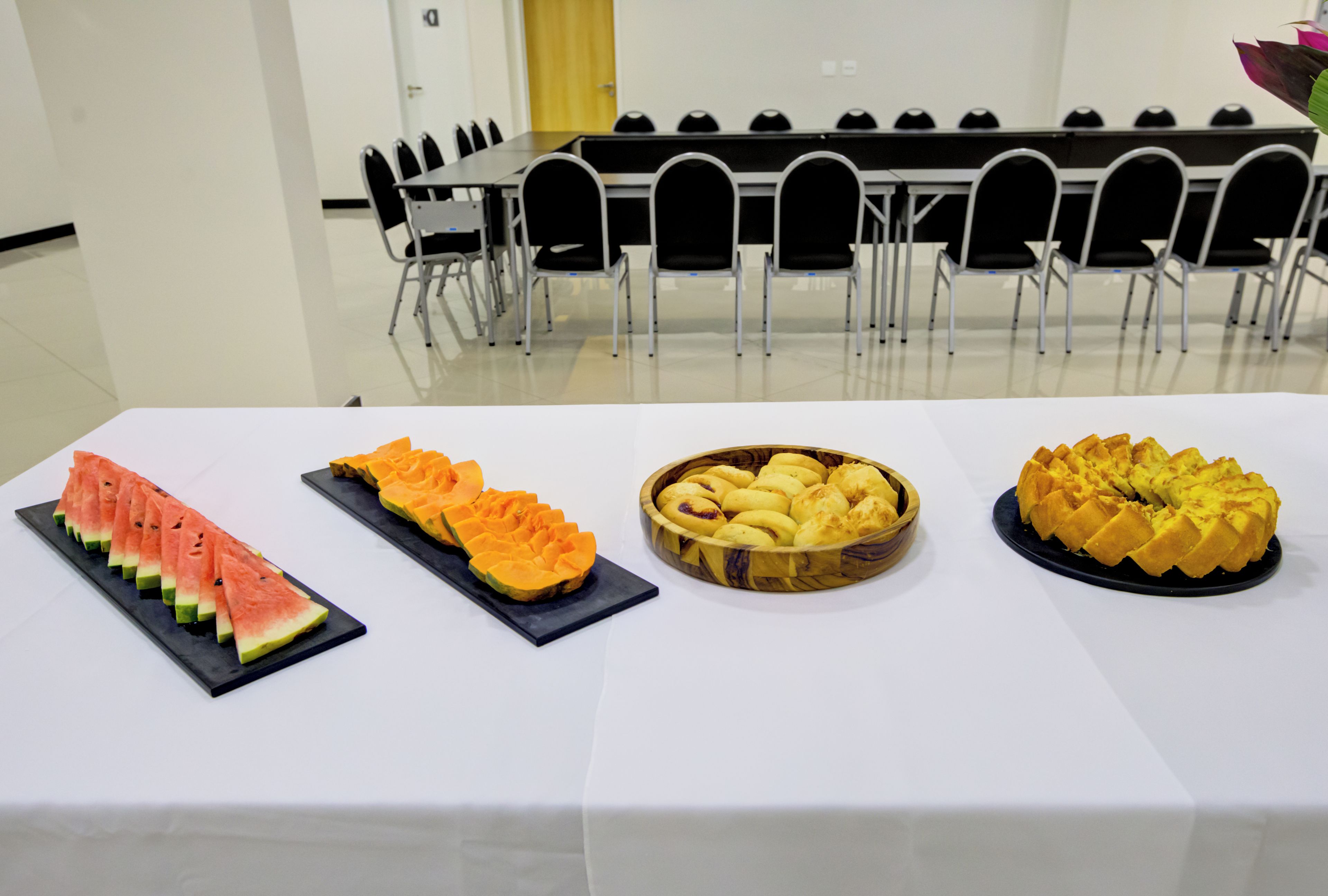 Sala de conferência com uma mesa comprida e cadeiras, mesa de buffet com frutas fatiadas e um bolo.